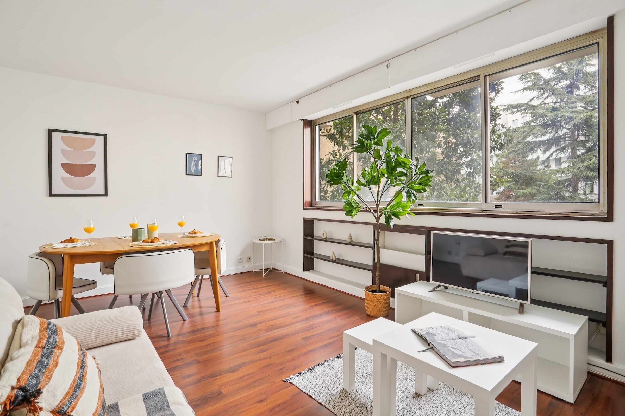 Uma sala de estar moderna com área de jantar, uma janela grande com vista para árvores, uma televisão de tela plana e um vaso de planta.
