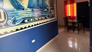 Quarto com um grande mural de veleiros, área de refeições com mesa e cadeiras e janela com cortinas vermelhas.