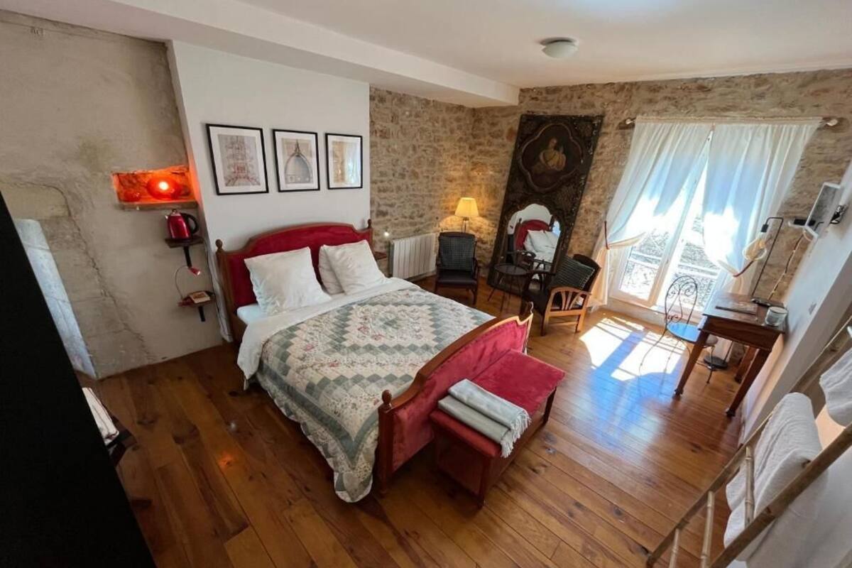 Quarto com piso de madeira, uma cama grande com cabeceira vermelha, uma mesinha, uma cadeira e uma janela com cortinas.