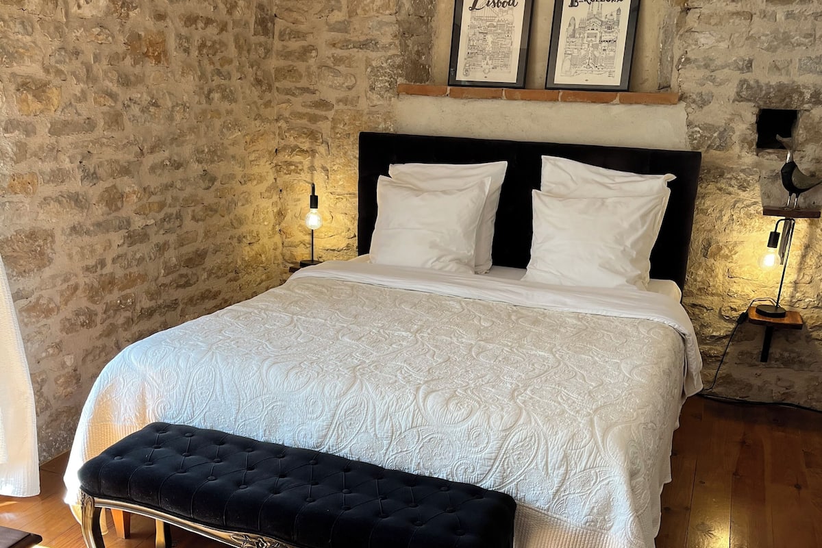 Quarto com parede de pedra, cama com roupa de cama branca, um banco preto e duas imagens emolduradas de Barcelona.