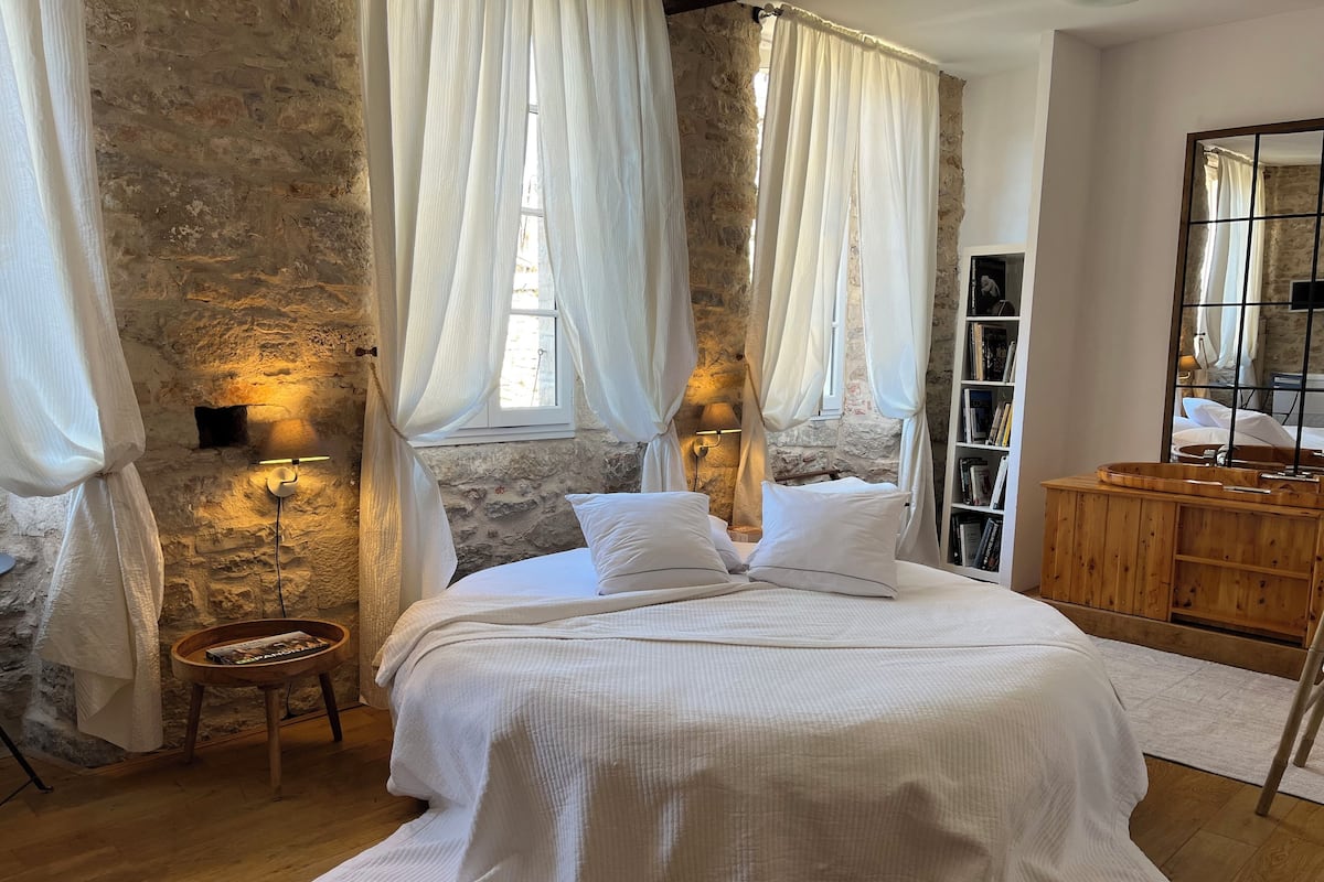 Quarto com parede de pedra, cama de madeira, cortinas brancas, abajur e penteadeira de madeira com espelho.