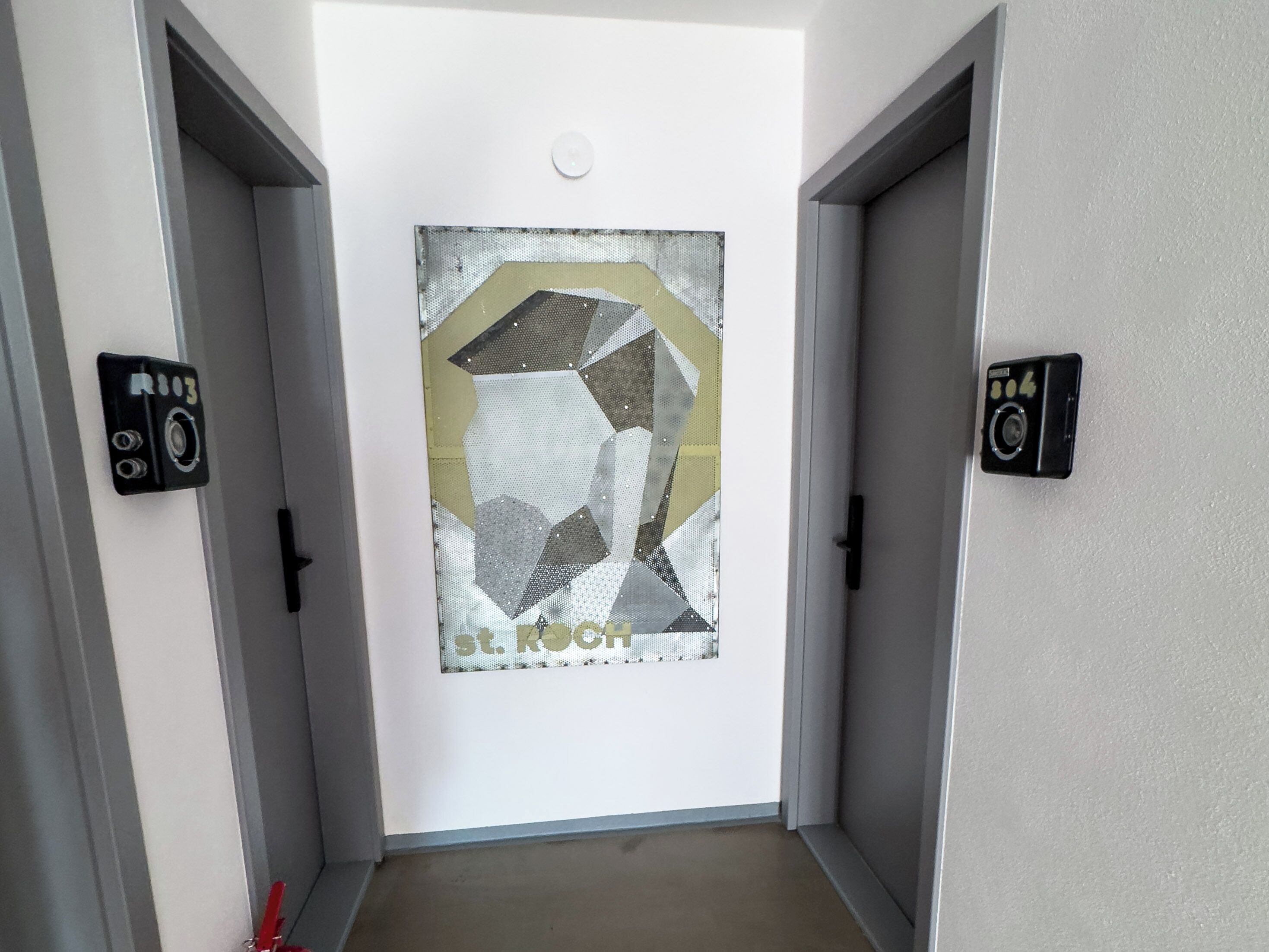 Um corredor com uma obra de arte geométrica na parede, dois interfones e um objeto vermelho no chão.
