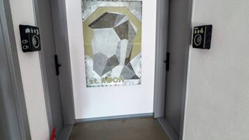 Um corredor com uma obra de arte geométrica na parede, dois interfones e um objeto vermelho no chão.
