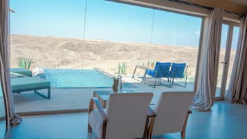 Quarto com uma grande janela com vista para uma paisagem desértica, piscina e área de estar com cadeiras.