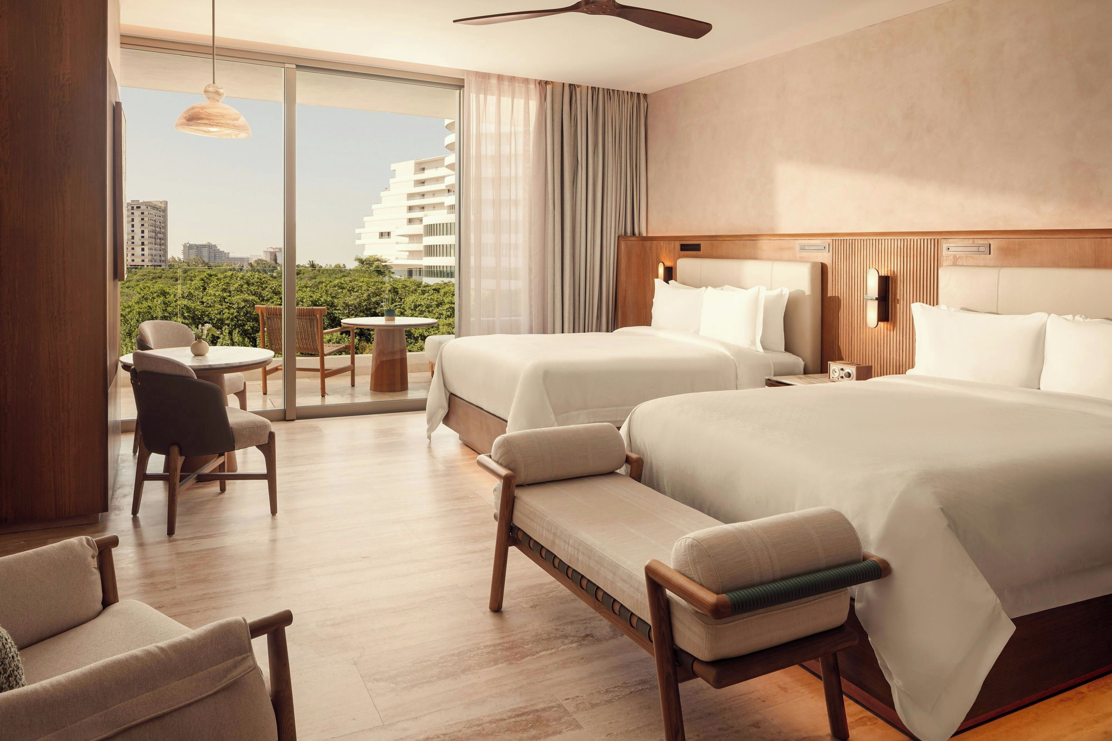 Quarto de hotel com duas camas, área de estar com mesa e varanda com vista para edifícios.