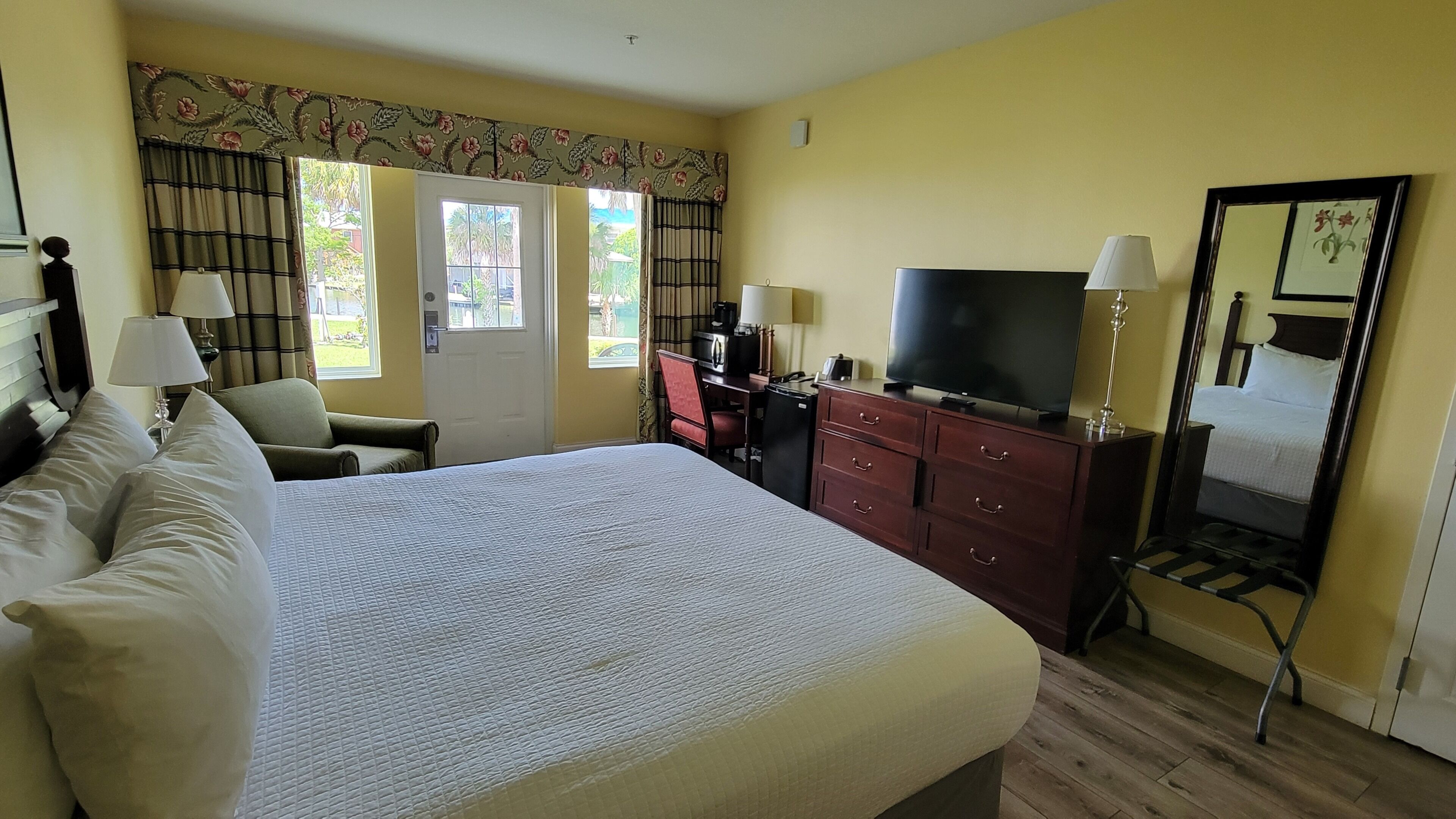 Quarto de hotel com uma cama grande, uma mesa de trabalho com televisão, cômoda e espelho.