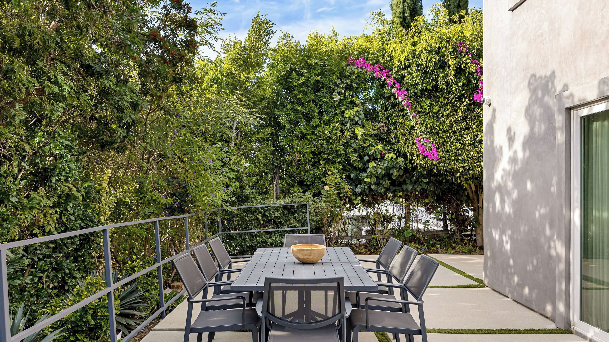 Um pátio externo com mesa e cadeiras, rodeado por vegetação exuberante e um edifício.