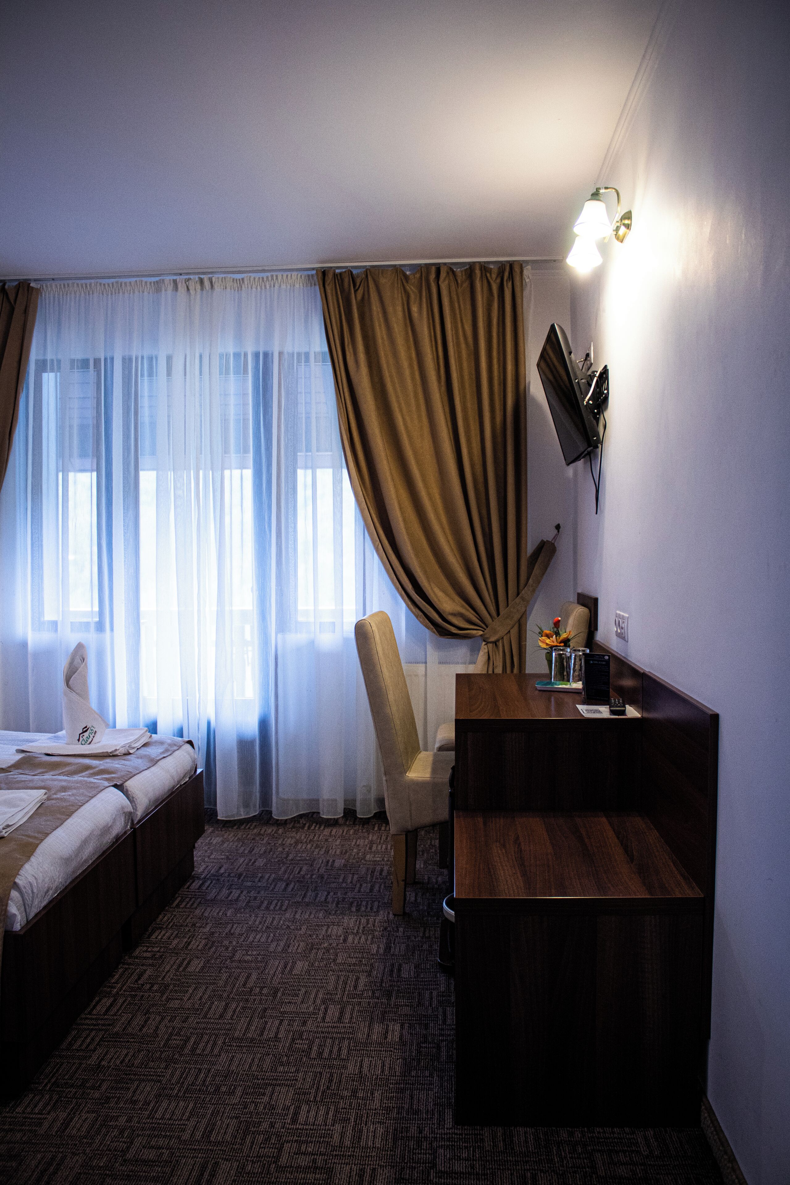 Quarto de hotel com cama, escrivaninha, cadeira, televisão e janela com cortinas.