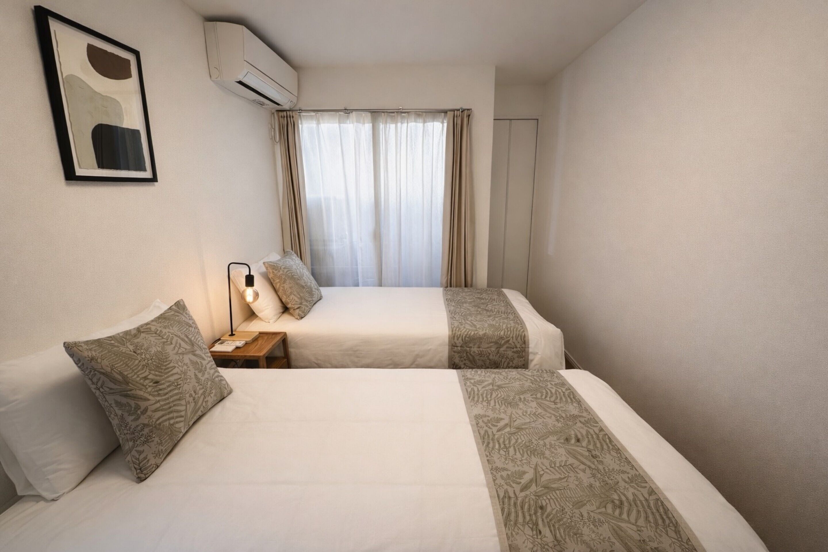 Quarto de hotel com duas camas, mesa de cabeceira, luminária, ar condicionado e janela com cortinas.