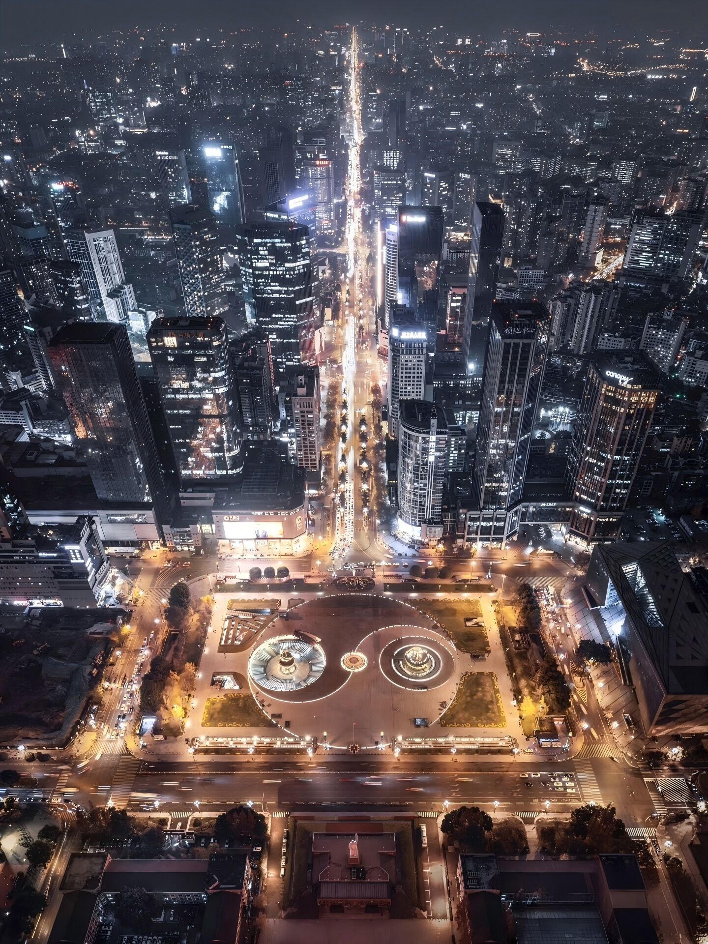 Vista aérea de uma paisagem urbana noturna com edifícios iluminados e uma praça central com padrões circulares.