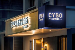 Placa CYBO Station com texto iluminado, fachada de um edifício moderno e porta de entrada visível.