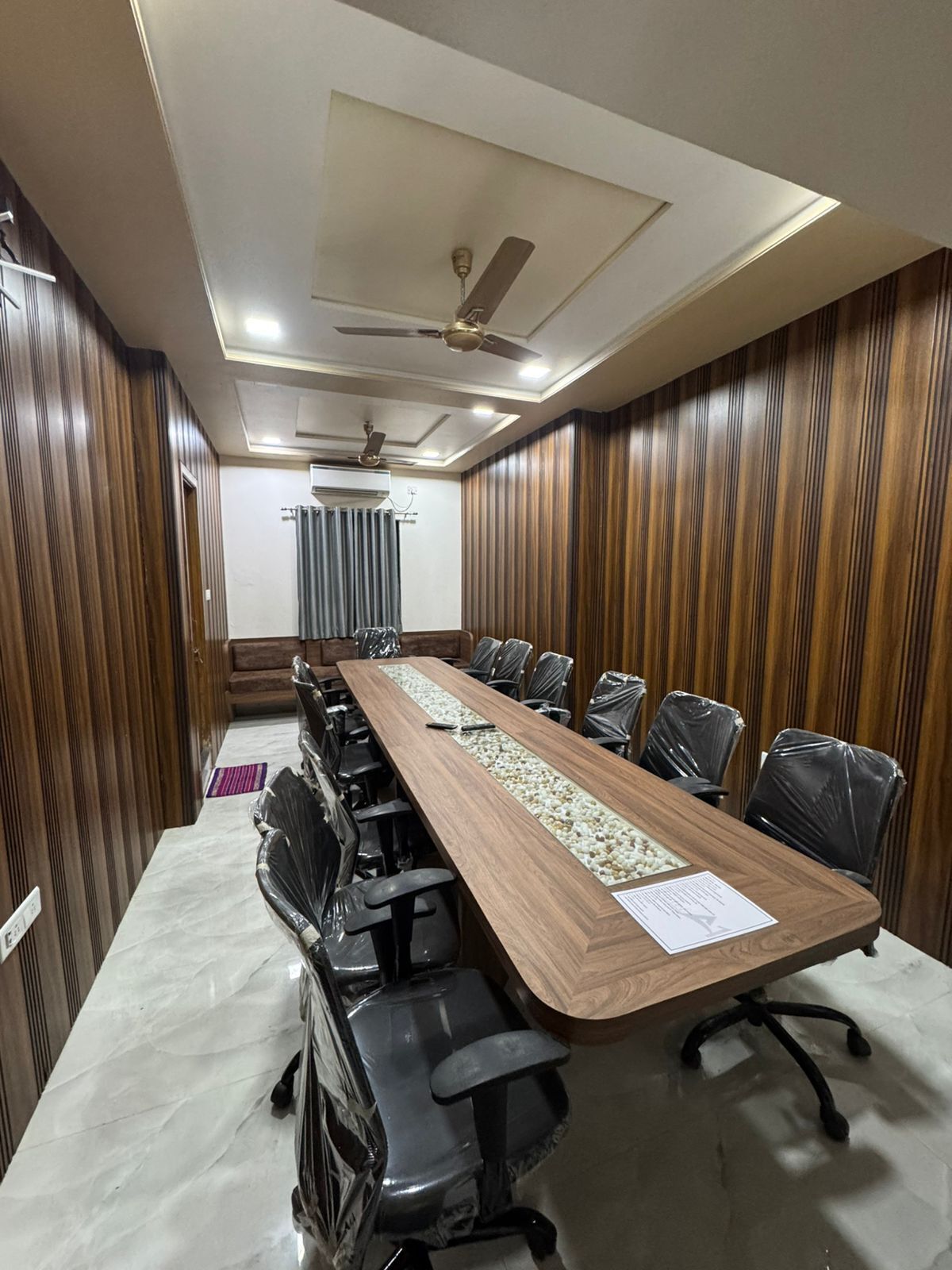 Uma sala de conferências com uma mesa de madeira comprida, cadeiras pretas, um ventilador de teto e lambris de madeira nas paredes.