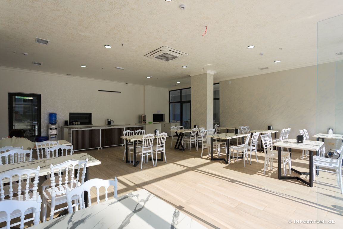 Uma ampla área de refeições interna com várias mesas e cadeiras, uma cozinha ao fundo e uma grande janela que proporciona luz natural.