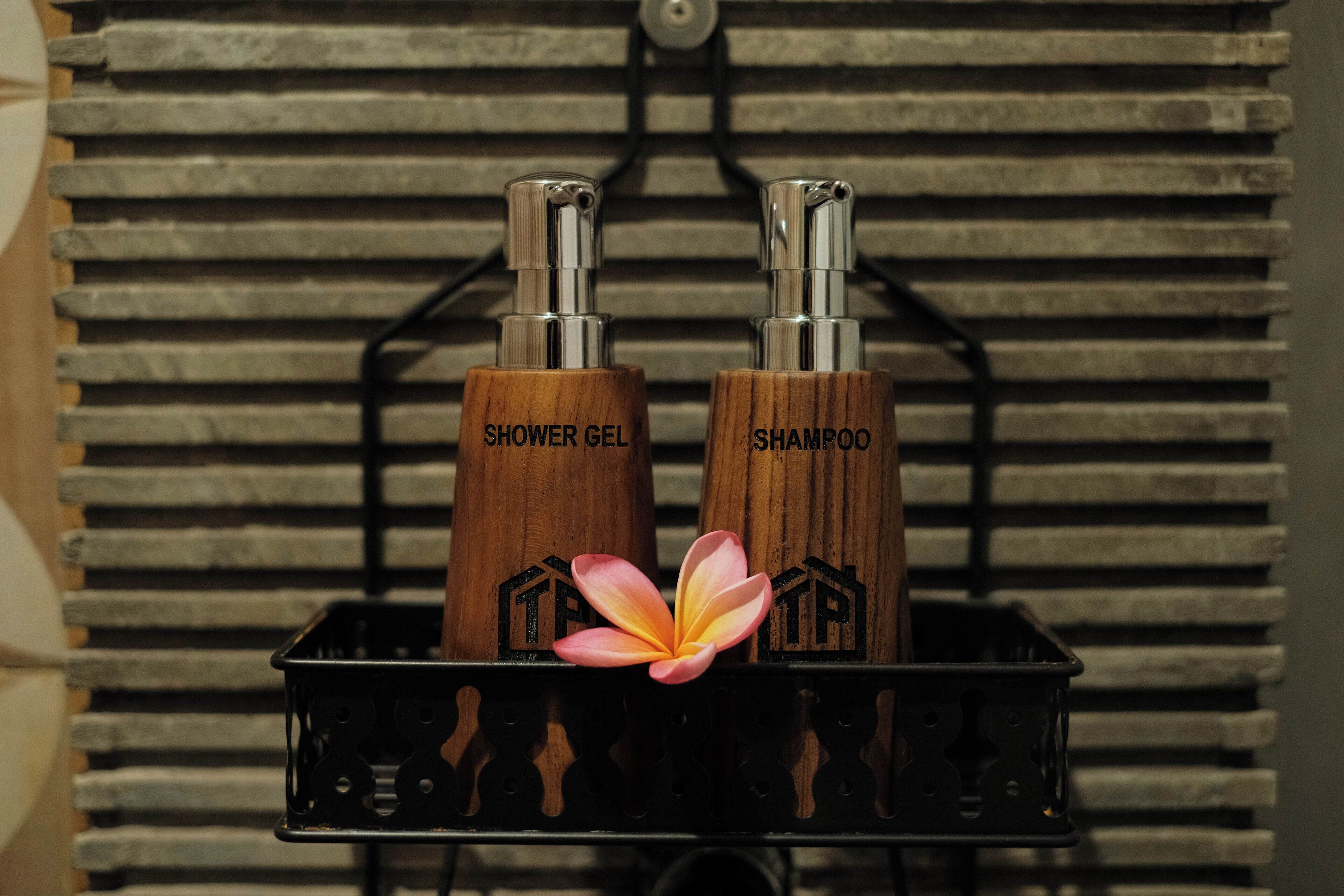 Duas garrafas de madeira com os dizeres 'gel de banho' e 'champô' numa bandeja preta com uma flor rosa.
