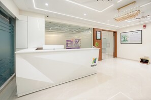 Uma área de recepção de hotel moderna, com um balcão de atendimento branco, um candelabro e um cartaz informativo emoldurado na parede.