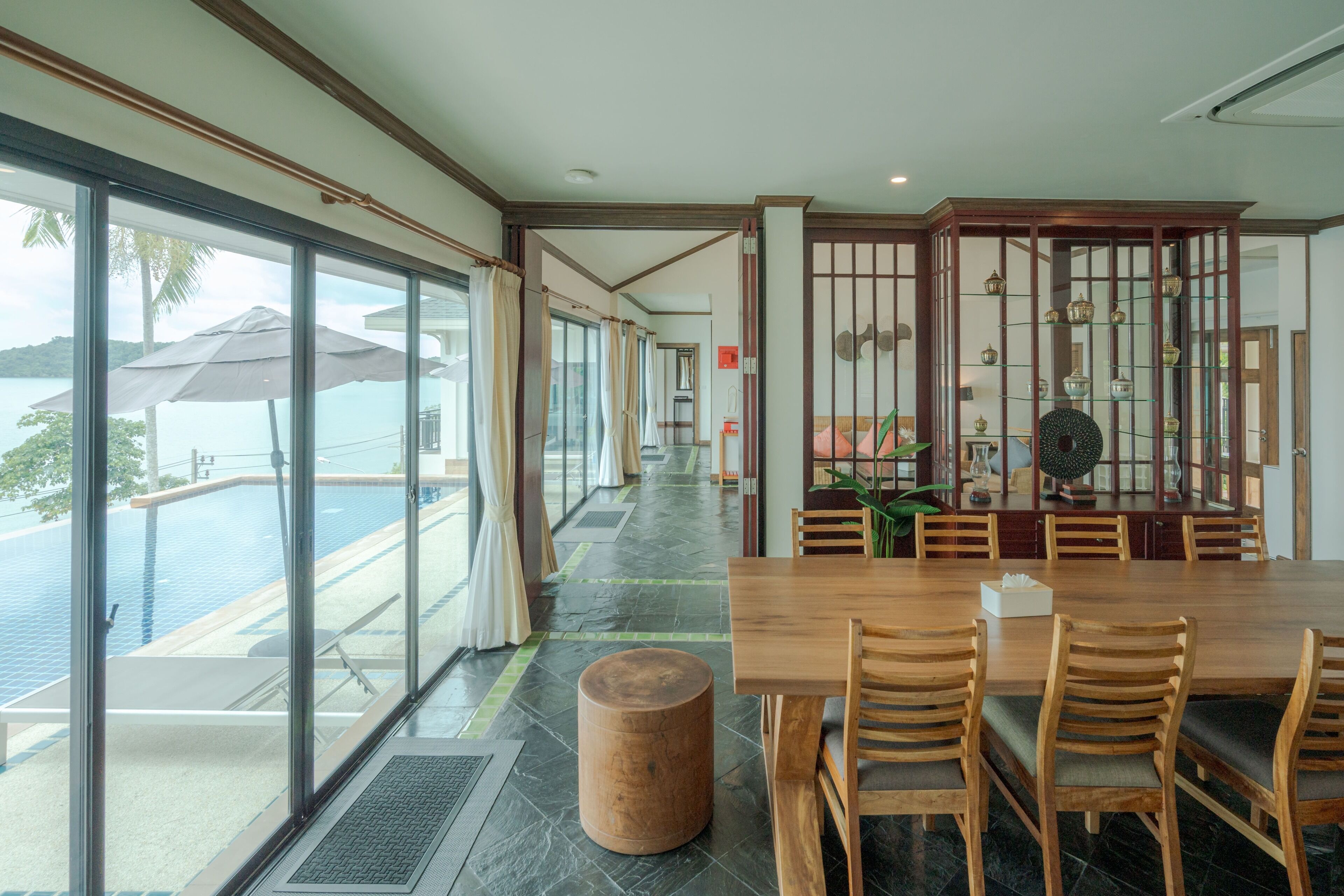 Uma sala de jantar ampla com uma grande mesa de madeira e cadeiras, um banquinho redondo pequeno e vista para uma piscina e palmeiras.