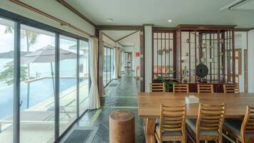 Uma sala de jantar ampla com uma grande mesa de madeira e cadeiras, um banquinho redondo pequeno e vista para uma piscina e palmeiras.