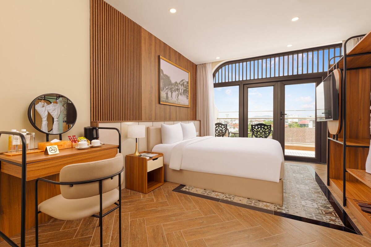 Quarto de hotel com cama, escrivaninha, cadeira, mesa de cabeceira e uma janela grande com vista.