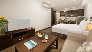 Quarto de hotel moderno com cama, televisão, mesa de jantar com café da manhã e uma cozinha compacta ao fundo.
