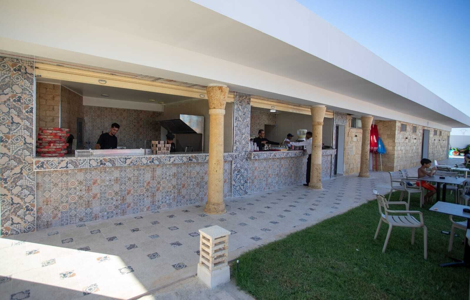 Uma área de bar ao ar livre com piso ladrilhado, balcão com revestimento de azulejos e espaço de estar com mesas e cadeiras.