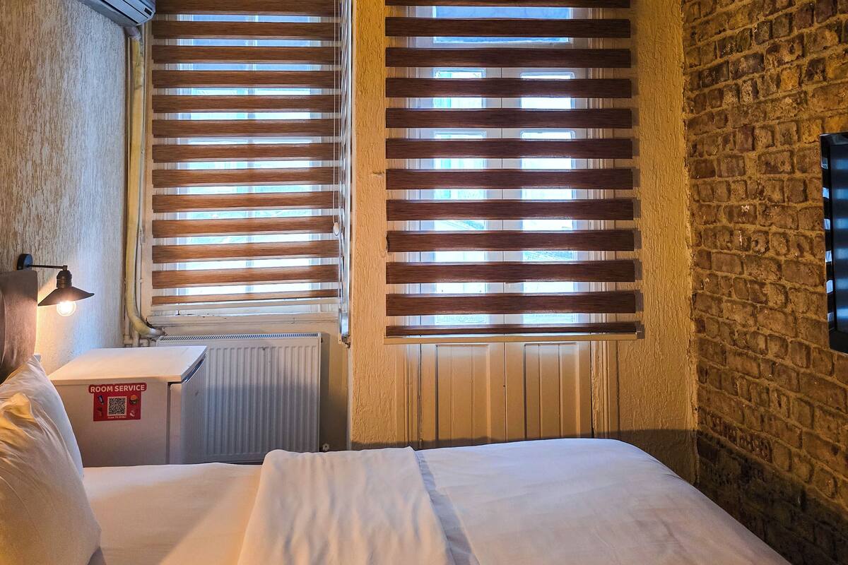 Quarto de hotel com cama, radiador, ar condicionado montado na parede e janela com persianas.