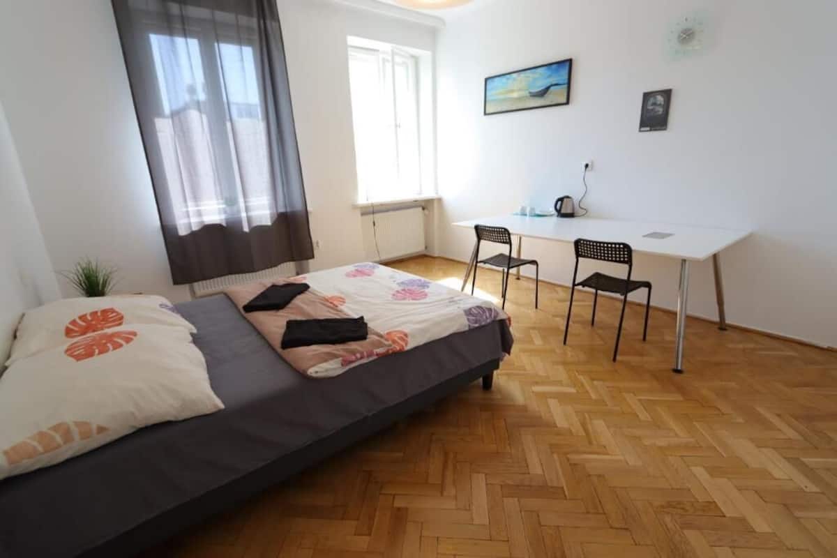 Quarto com cama, escrivaninha e cadeira. Há uma janela com cortinas, um radiador e uma pequena planta.