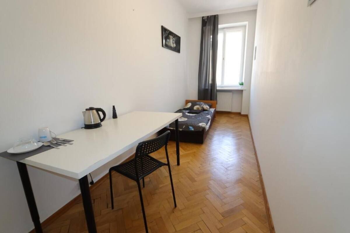 Quarto pequeno e limpo com uma mesa branca, uma cadeira preta, uma cama com manta cinza e branca, e uma janela com cortinas.