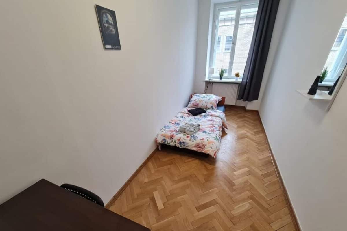 Um quarto pequeno e estreito com piso de madeira em padrão herringbone, uma cama simples com roupa de cama florida, uma mesa pequena e uma janela com cortinas.