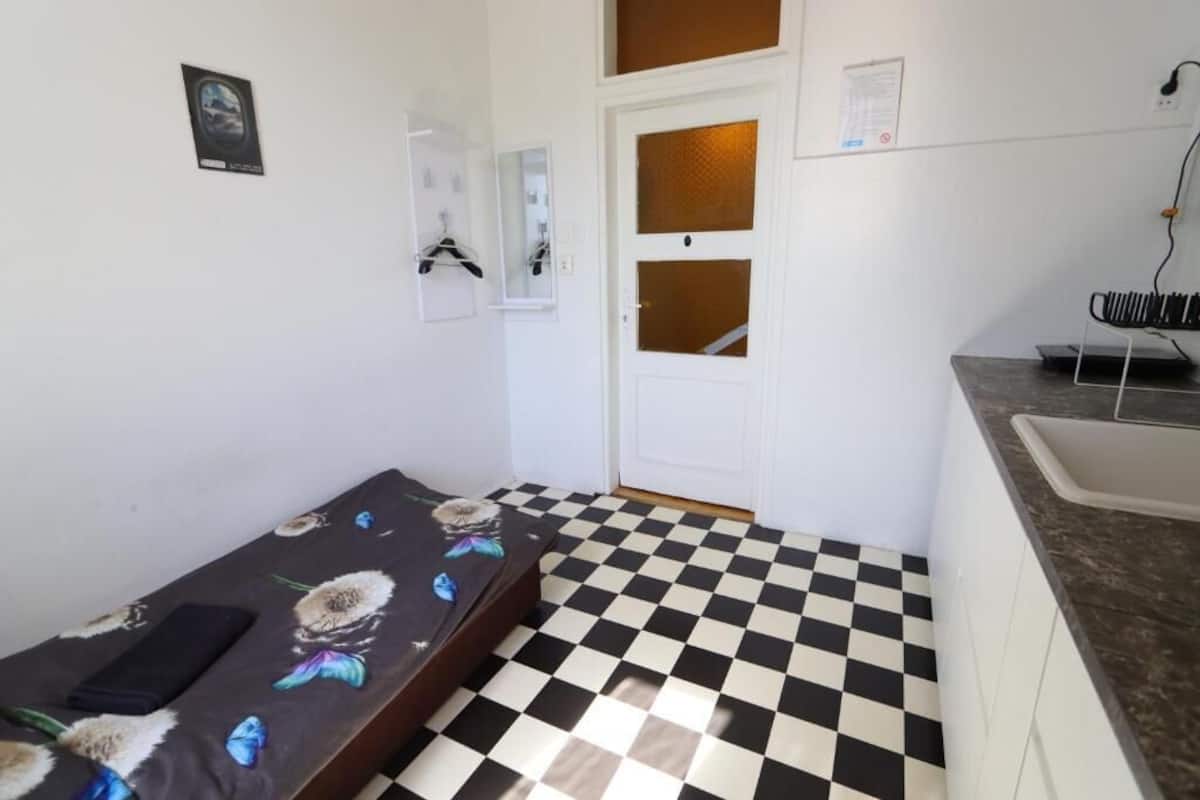 Um quarto pequeno com piso xadrez, uma cama com colcha florida, uma porta branca, uma janela pequena e uma área de cozinha compacta.