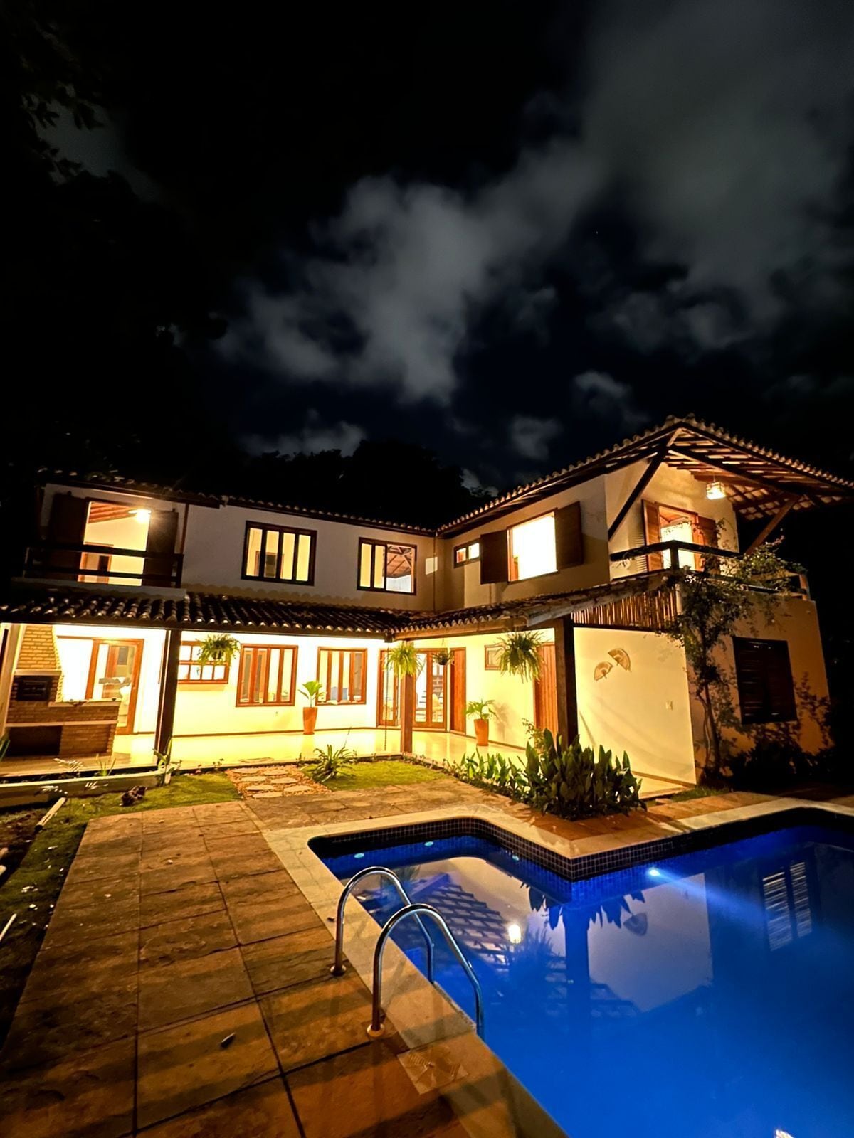 Uma casa de dois andares com piscina, um pátio e fachada bem iluminada à noite.