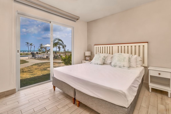 Quarto com uma cama grande, uma mesa de cabeceira e vista para uma praia e palmeiras através de uma porta de correr de vidro.