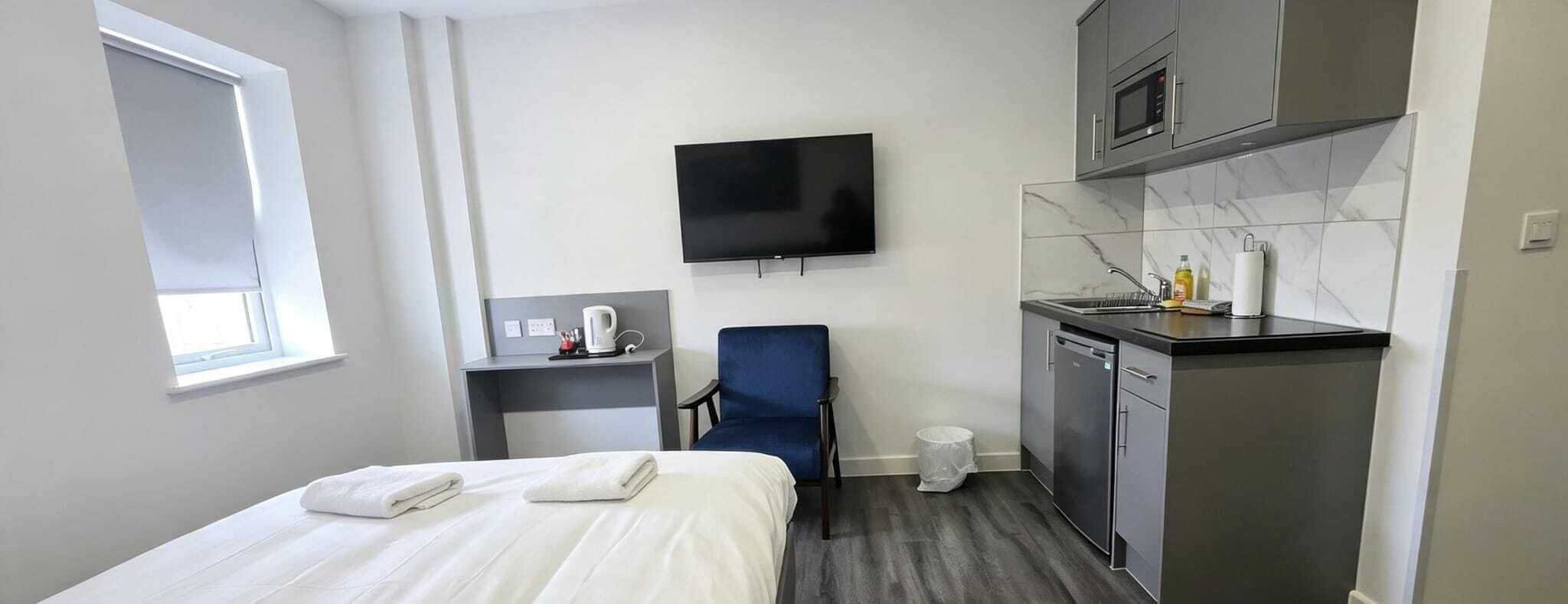 Quarto de hotel compacto com cama, pequena escrivaninha, cadeira, televisão e cozinha compacta.