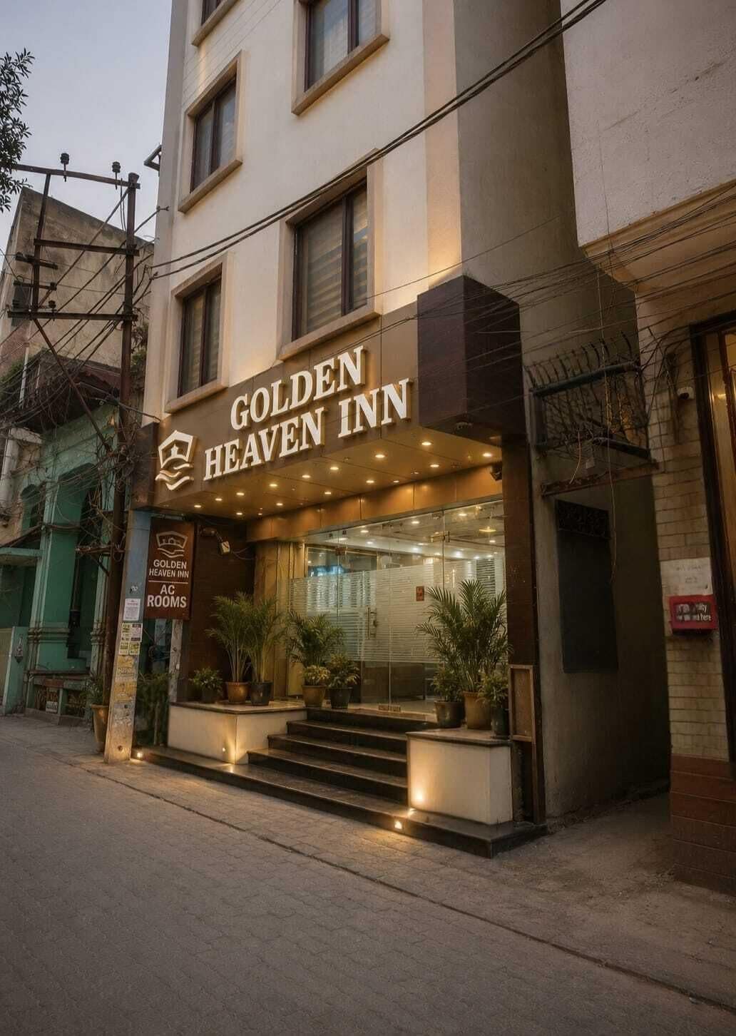 Exterior do Golden Heaven Inn, com entrada notável e degraus que levam à porta, vasos de plantas e vista clara do interior através da fachada de vidro.
