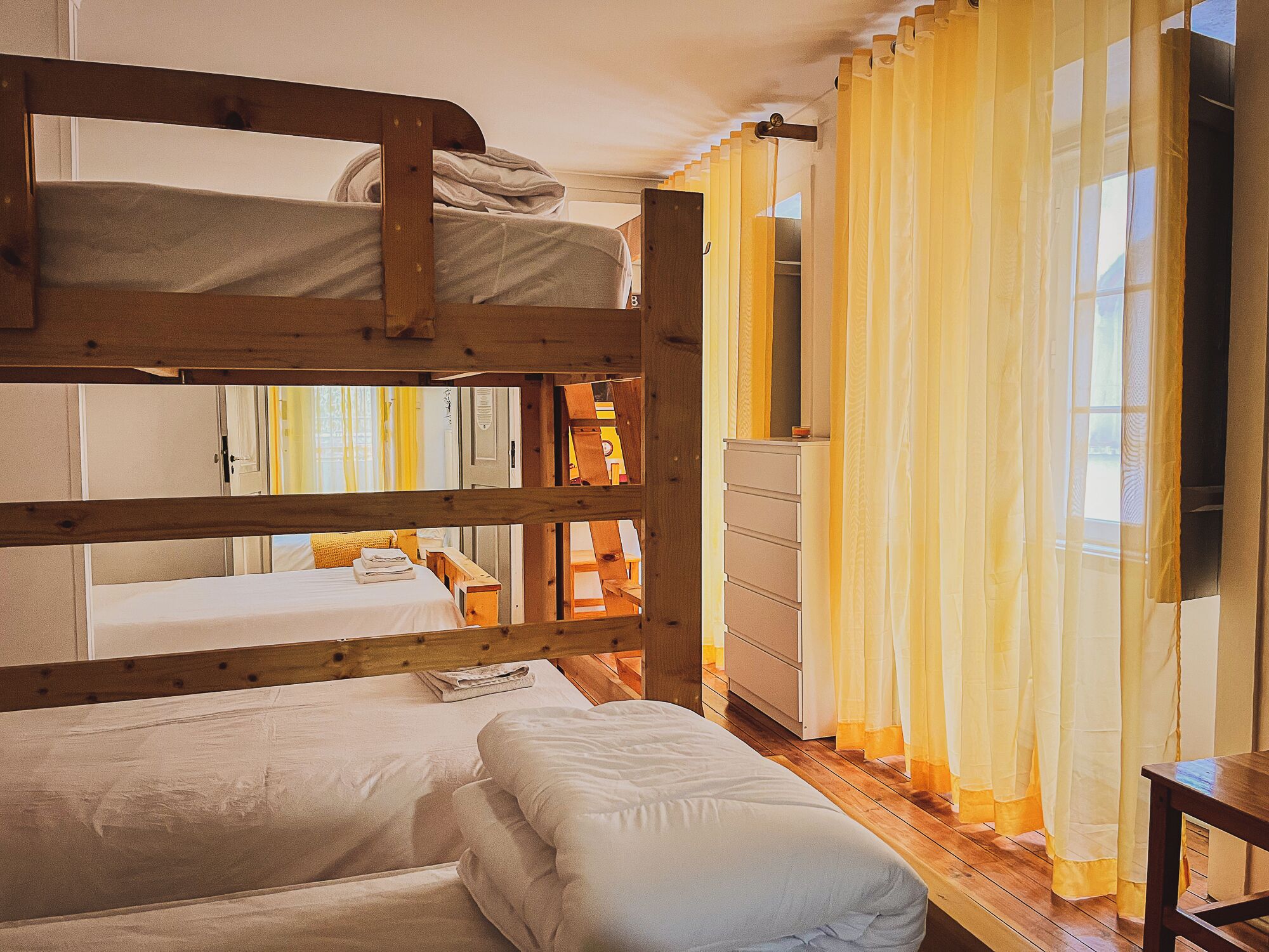Quarto com beliche, cabeceira de madeira, uma mesinha e janela com cortinas amarelas.