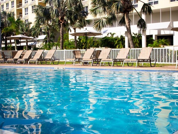 Piscina com espreguiçadeiras e guarda-sóis, cercada por palmeiras e um edifício de vários andares.