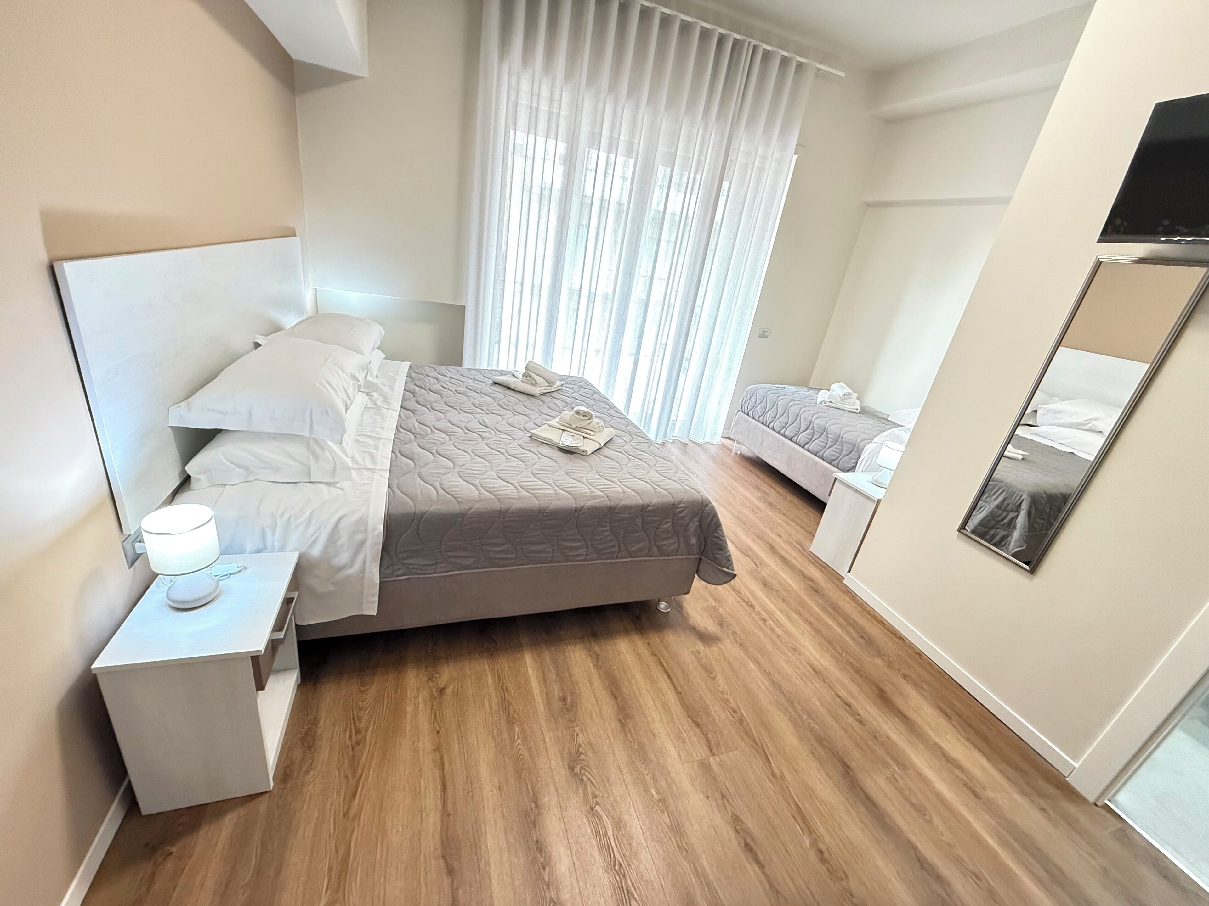 Quarto de hotel moderno com uma cama grande, mesa de cabeceira, espelho e janela com cortinas translúcidas.