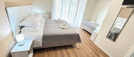 Quarto de hotel moderno com uma cama grande, mesa de cabeceira, espelho e janela com cortinas translúcidas.
