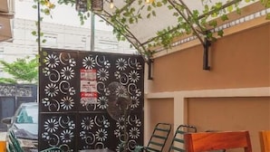 Uma área externa com assentos de metal verde e uma mesa de madeira, sob uma estrutura coberta com luzes pendentes e folhagem.