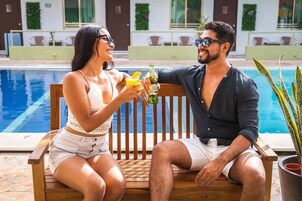 Um casal sentado à beira da piscina com bebidas, um banco de madeira e um vaso de planta.