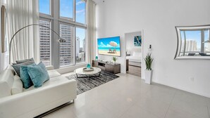 Uma sala de estar moderna com um sofá branco, uma mesa de centro e uma televisão grande exibindo uma cena de praia.