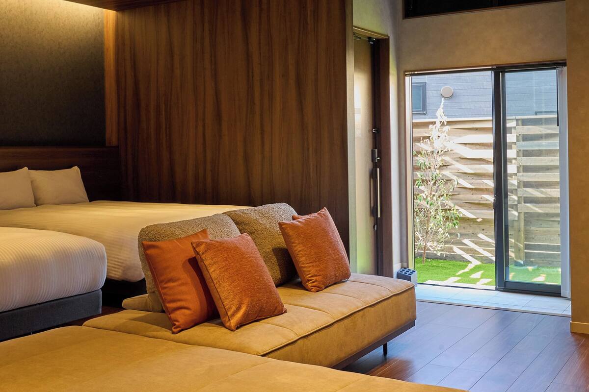 Quarto de hotel com duas camas, um sofá e uma porta de vidro que desliza para o exterior.