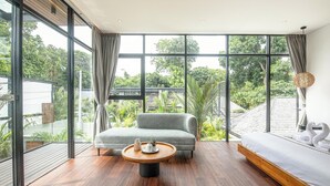 Quarto moderno com uma janela grande, um sofá cinzento, uma mesa de centro de madeira redonda e uma cama com roupa de cama branca.