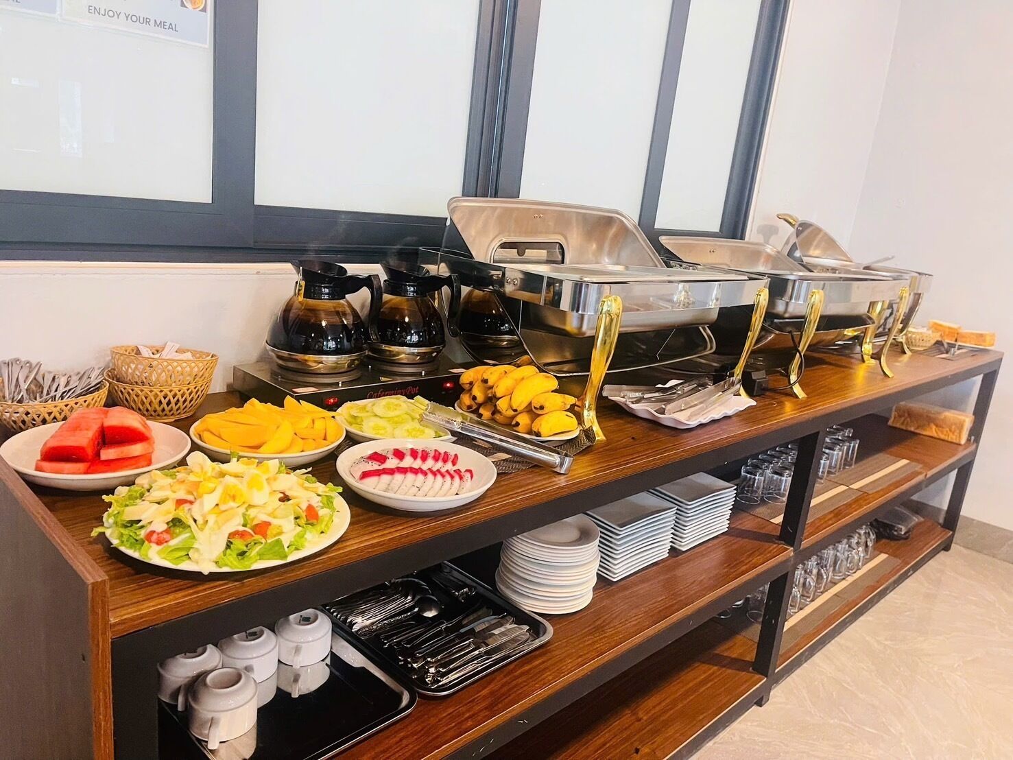 Disposição de buffet com diversos alimentos, incluindo frutas, saladas e bebidas, dispostos sobre uma mesa de madeira com estação de café e dispensadores de condimentos.