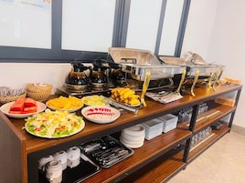 Disposição de buffet com diversos alimentos, incluindo frutas, saladas e bebidas, dispostos sobre uma mesa de madeira com estação de café e dispensadores de condimentos.