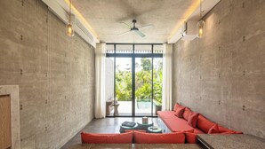 Um interior moderno com parede de concreto, sofá vermelho, ventilador de teto e vista para área externa através de grandes portas de vidro.