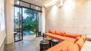 Uma sala de estar moderna com um sofá grande laranja, uma porta de vidro que dá para uma piscina e uma parede de concreto com um aparelho de iluminação.