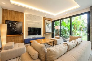 Uma sala de estar moderna com um sofá grande, uma mesa de centro e uma TV de tela plana. Há quadros nas paredes e vista para o verde lá fora.