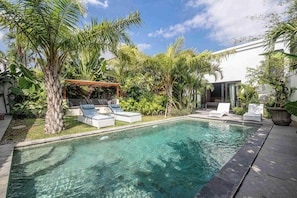 Piscina com espreguiçadeiras, cercada por plantas tropicais e uma pérgula.