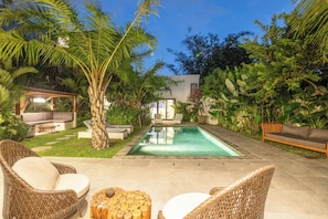 Uma área à beira da piscina com móveis de vime, uma mesa de madeira e vegetação exuberante.