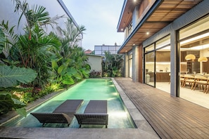 Uma área moderna com piscina ao ar livre, duas espreguiçadeiras de vime, rodeada por plantas tropicais exuberantes e um deck de madeira.